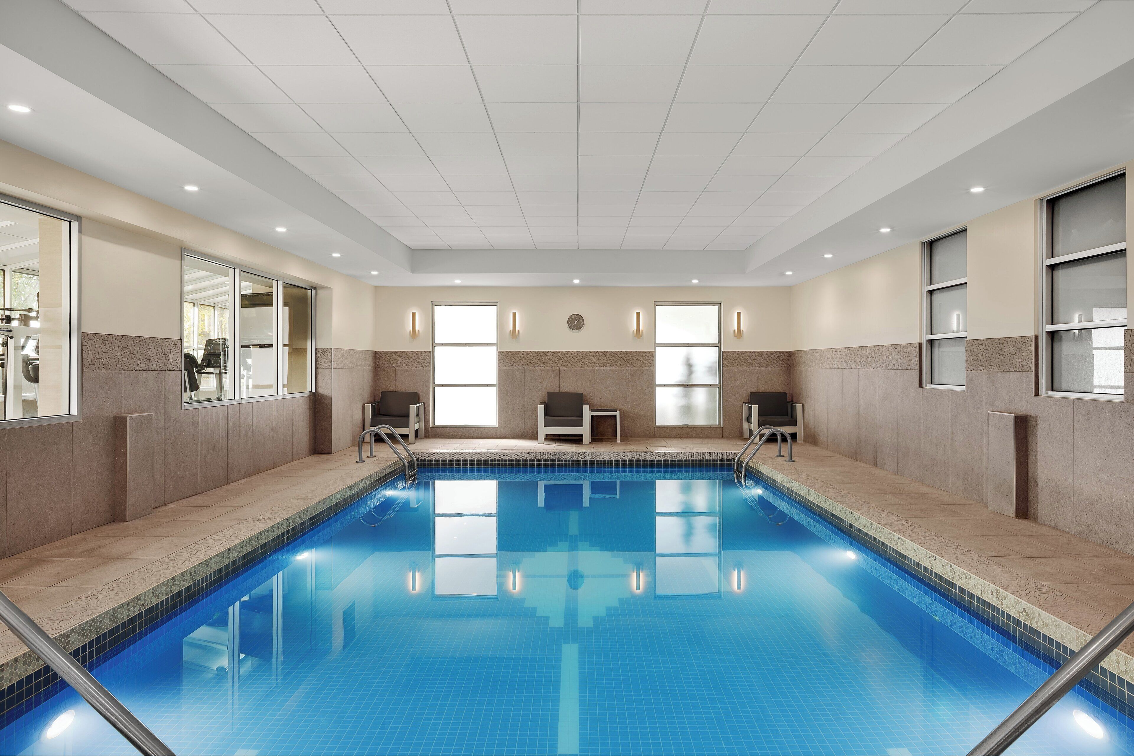 Piscina interior com água azul cristalina, rodeada por paredes bege e chão ladrilhado. Há duas cadeiras e uma pequena mesa perto da piscina.
