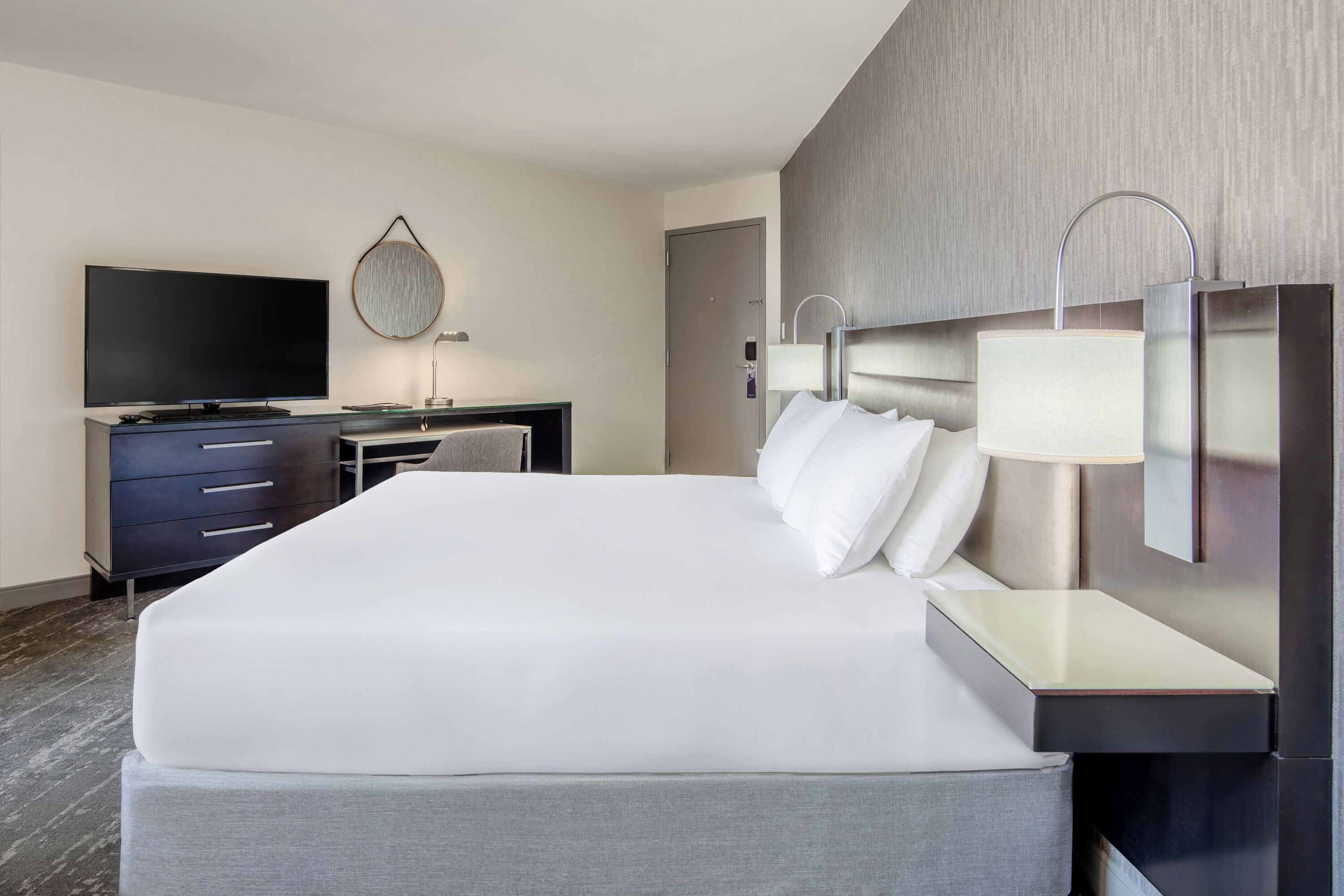 Quarto de hotel moderno com uma cama grande, televisão de tela plana, escrivaninha com cadeira e luminária na parede.