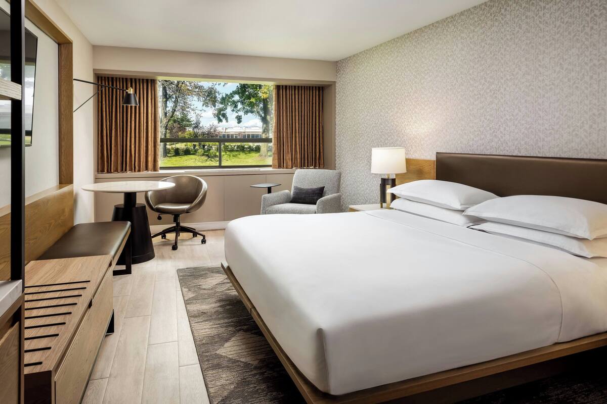 Quarto de hotel com uma cama grande, uma escrivaninha com cadeira, uma mesinha e vista para o exterior.