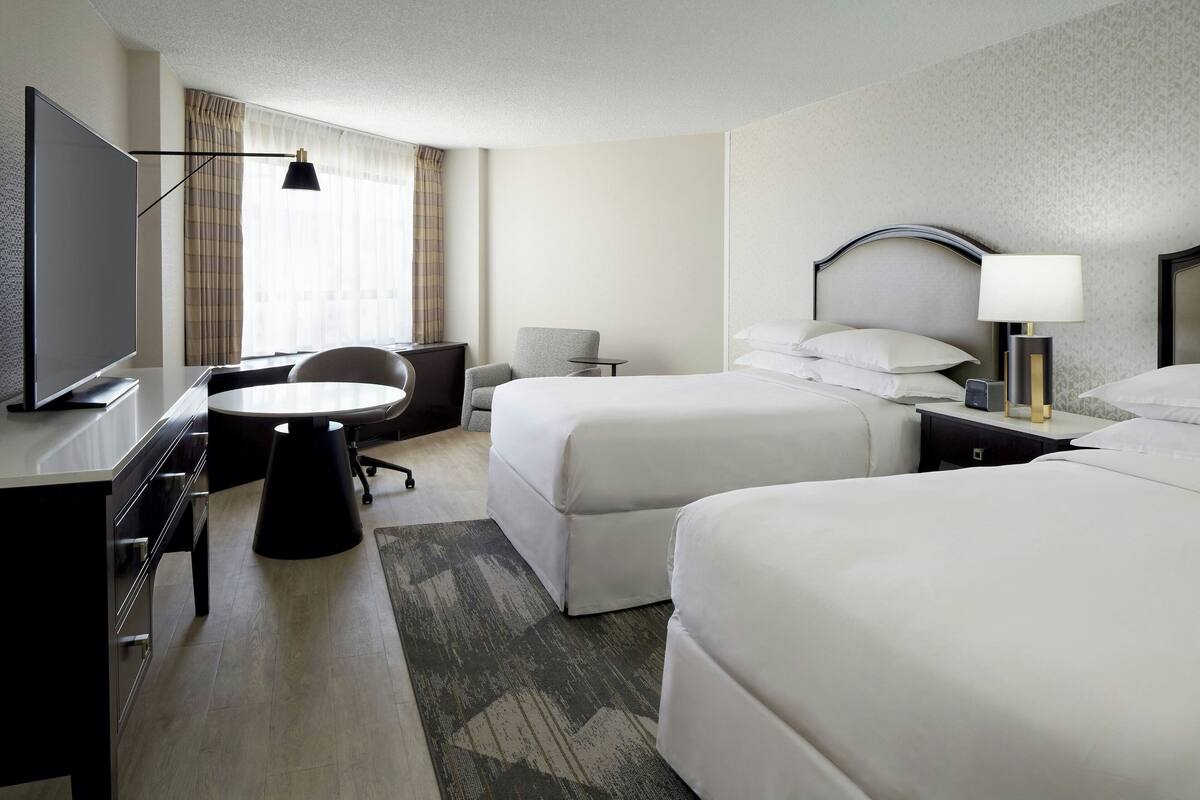 Quarto de hotel com uma cama grande, uma escrivaninha com televisão, uma cadeira e uma mesa pequena.