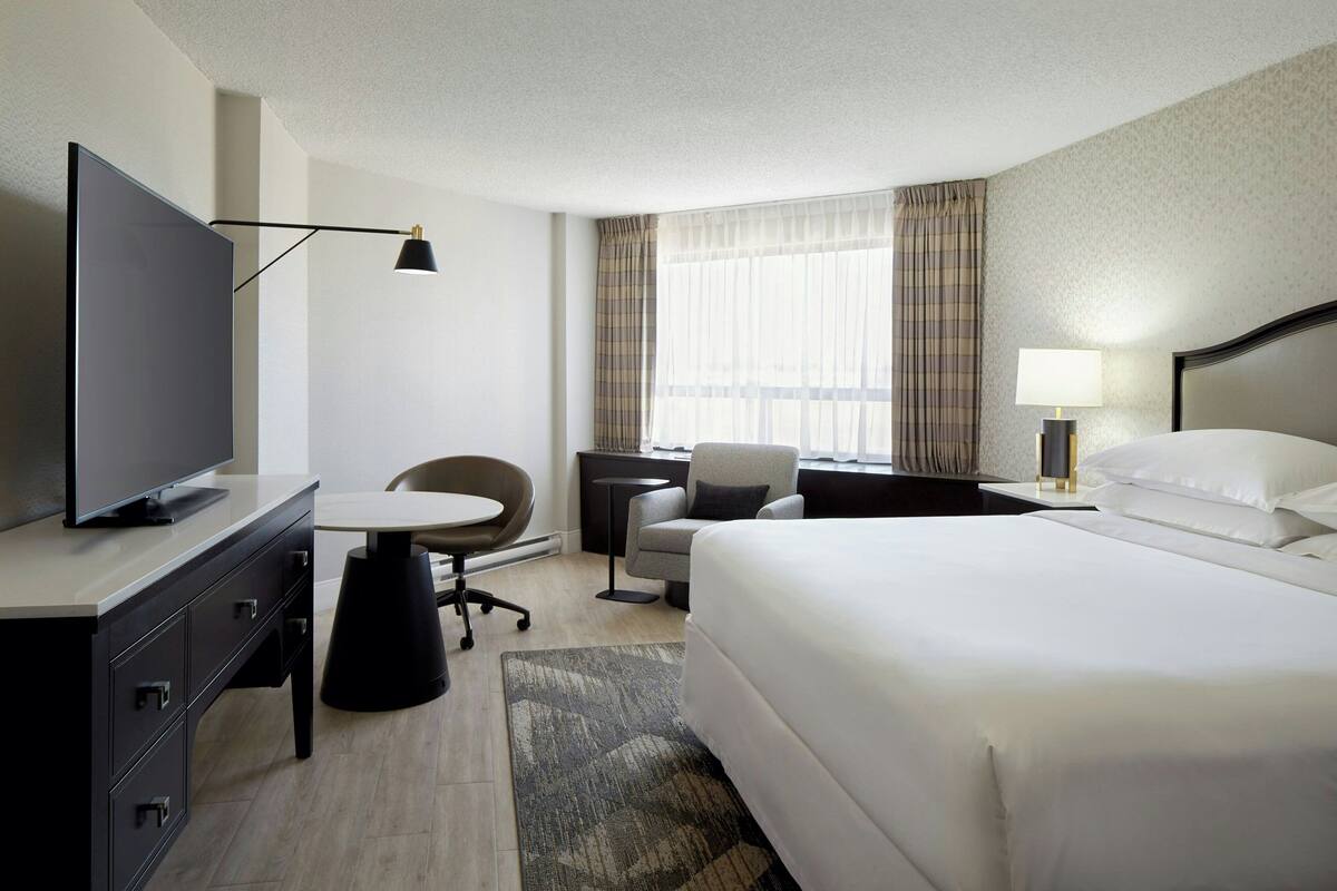 Quarto de hotel com cama, escrivaninha com televisão, cadeira, mesa pequena e abajur.