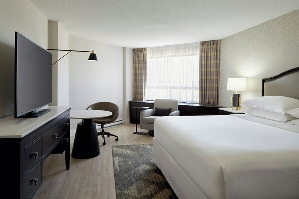 Quarto de hotel com cama, escrivaninha com televisão, cadeira, mesa pequena e abajur.