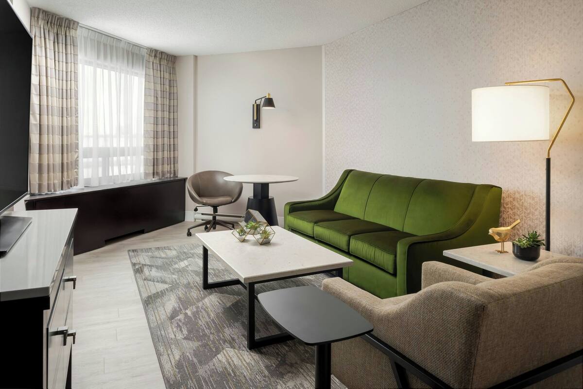 Uma sala de estar moderna com um sofá verde, uma mesa de centro branca, uma mesinha lateral, uma televisão e um abajur.