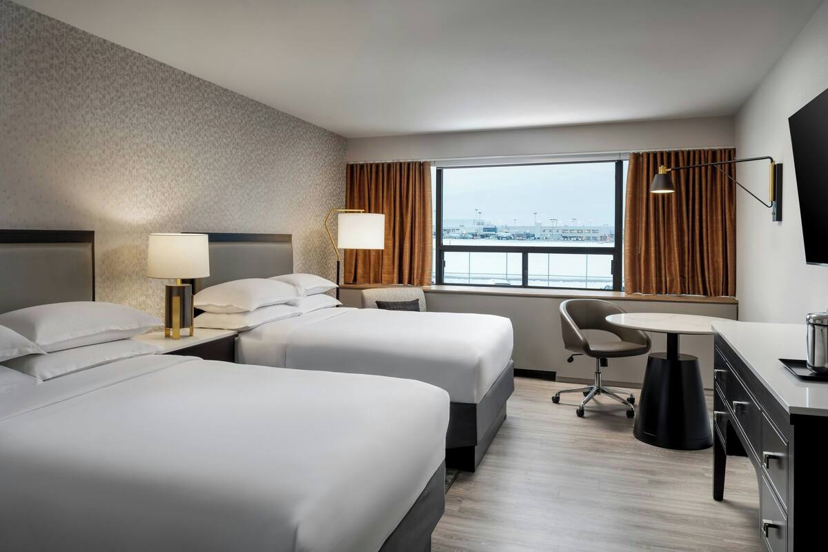 Quarto de hotel com duas camas, uma escrivaninha, uma cadeira, uma televisão e uma janela com cortinas.