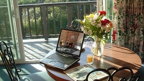 Um laptop sobre uma mesa, com um jornal, um vaso de flores e vista para árvores através de uma janela.