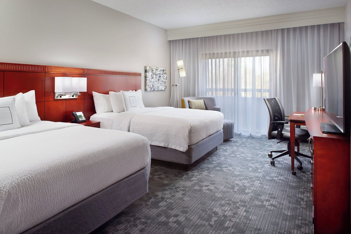 Quarto de hotel com duas camas, uma escrivaninha, uma cadeira, uma televisão e uma janela grande com cortinas.