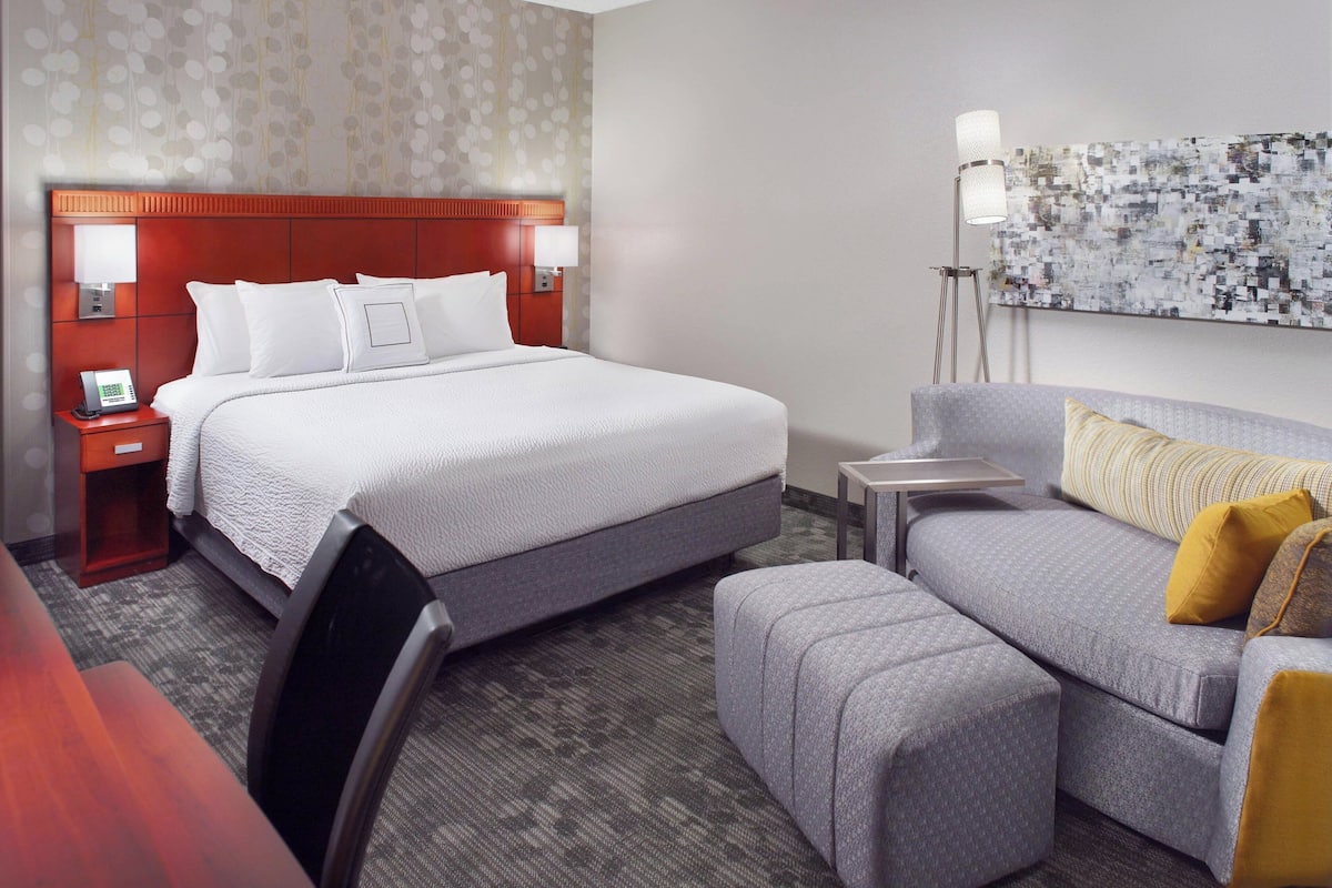 Quarto de hotel com uma cama grande, um sofá cinzento, uma mesinha e um abajur.