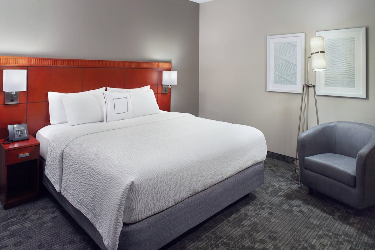 Quarto de hotel com uma cama grande, uma mesa de cabeceira com telefone, duas luminárias na parede e uma poltrona cinza.