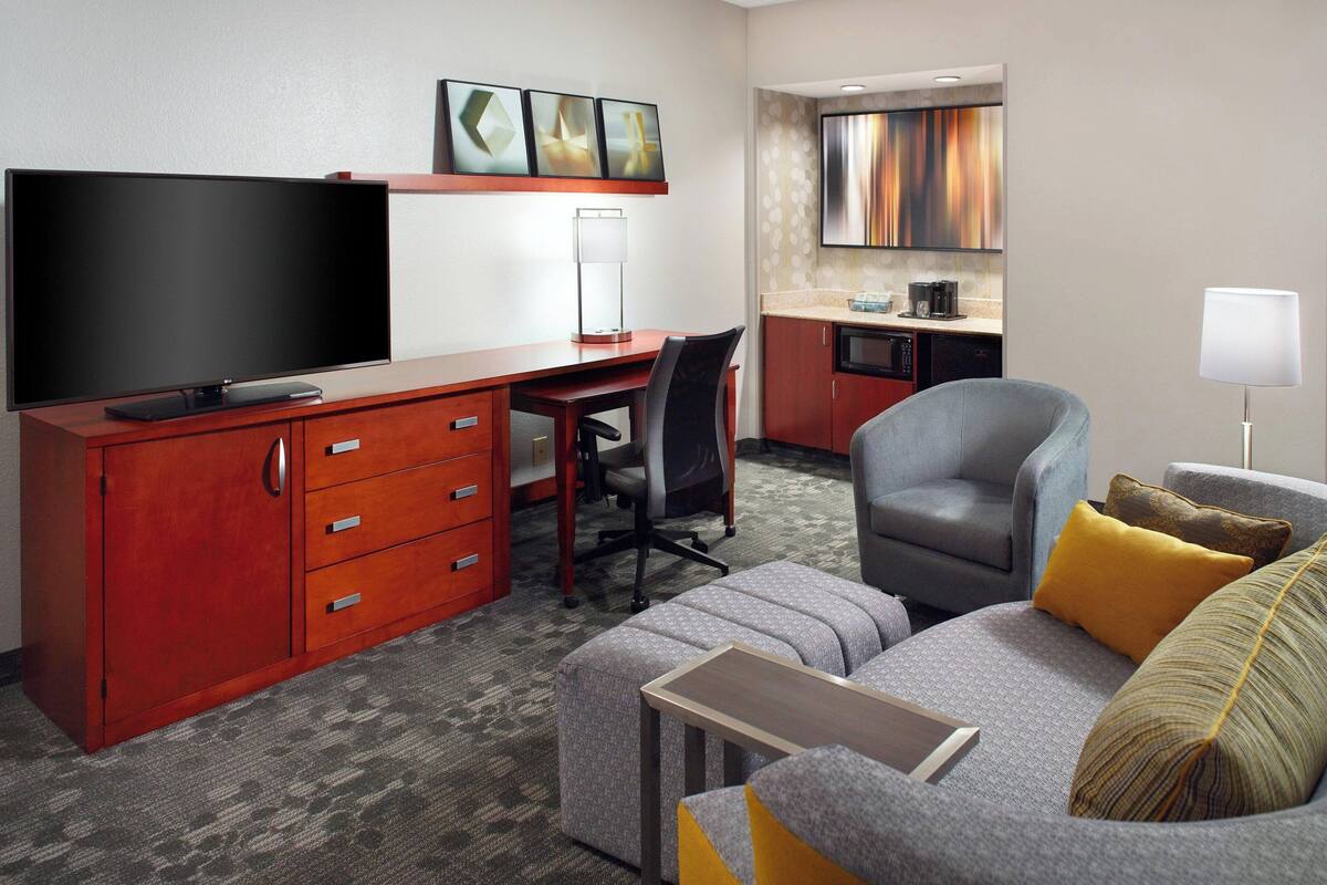 Quarto de hotel com televisão de ecrã plano, secretária com cadeira, sofá, mesa pequena e cozinha compacta.