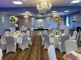 Uma sala de banquetes com mesas redondas preparadas para um evento formal, com cadeiras brancas, louças douradas e arranjos florais.