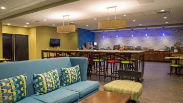 Um hall de hotel moderno com um sofá azul, uma mesa de centro de madeira, área de bar com banquetas e um fundo de parede de pedra.