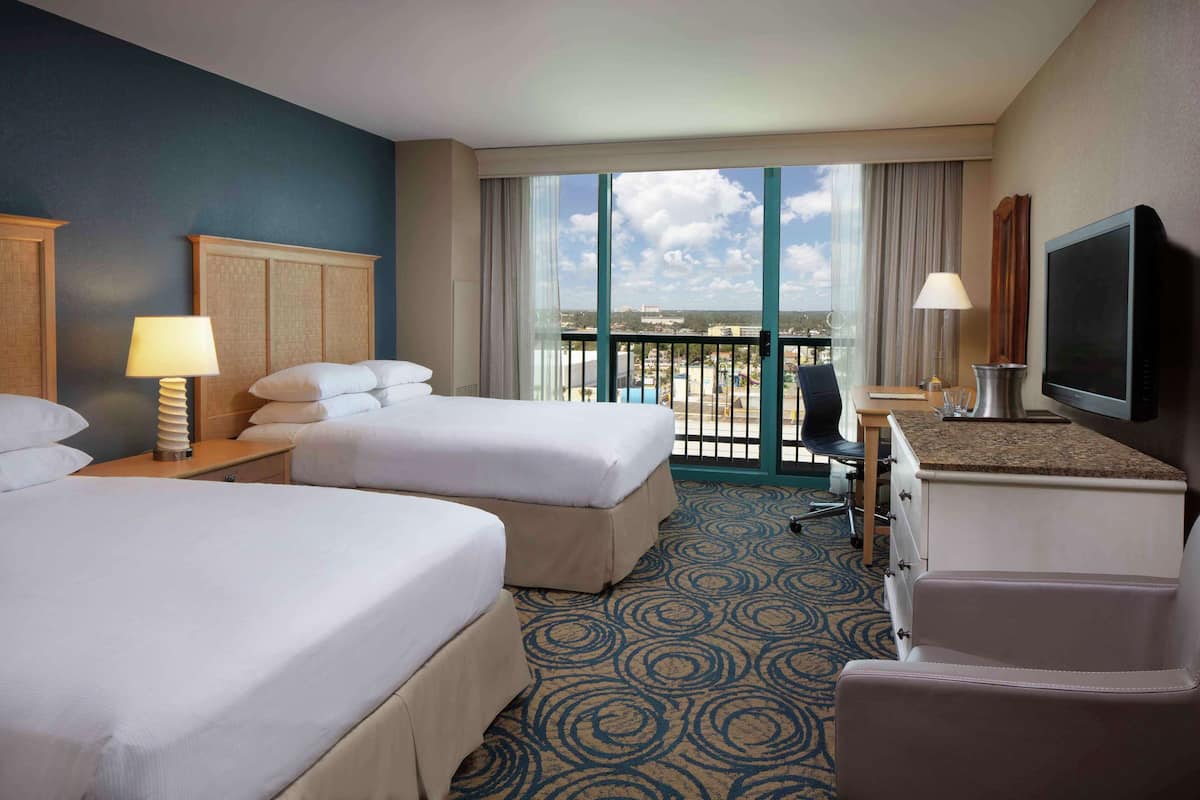Quarto de hotel com duas camas, uma mesa, uma televisão e uma varanda com vista.