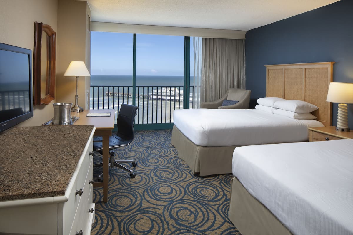 Quarto de hotel com uma cama grande, uma cama menor, uma escrivaninha com luminária, uma cadeira e vista para o oceano.