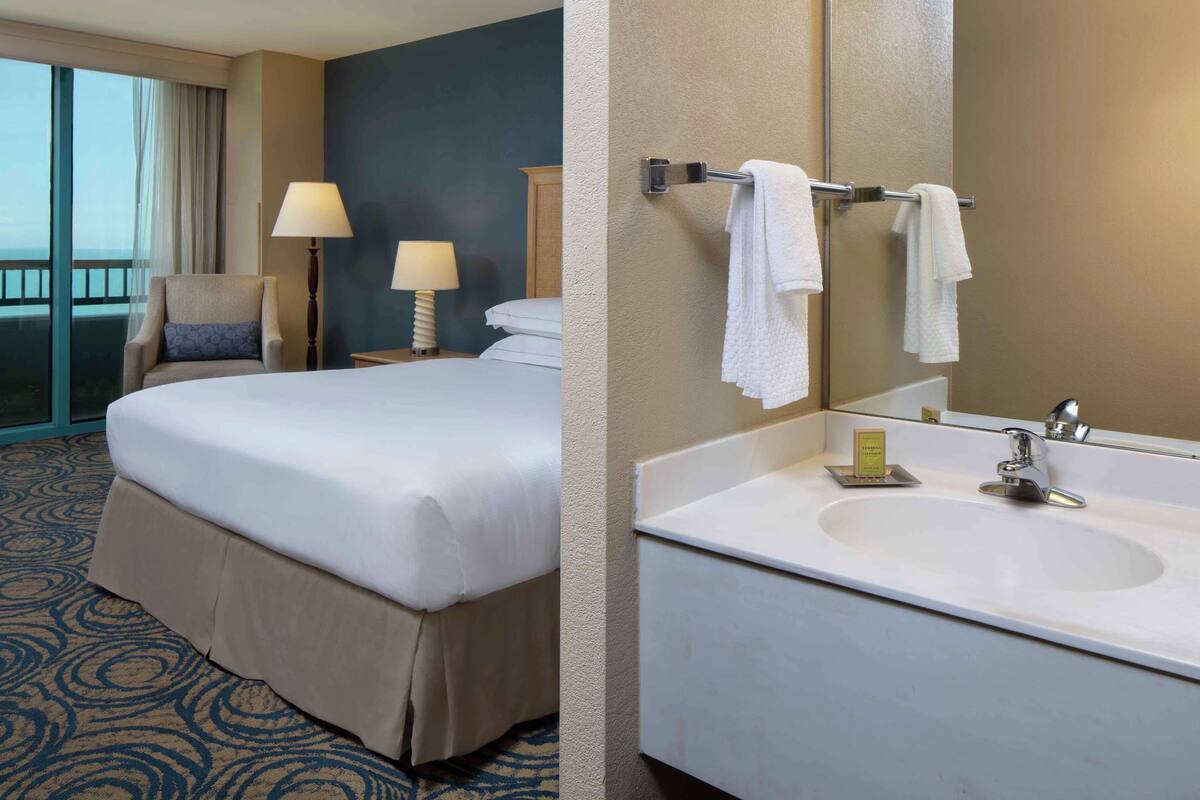 Quarto de hotel com uma cama grande, uma cadeira, duas luminárias de cabeceira, banheiro com pia e espelho e um porta-toalhas.