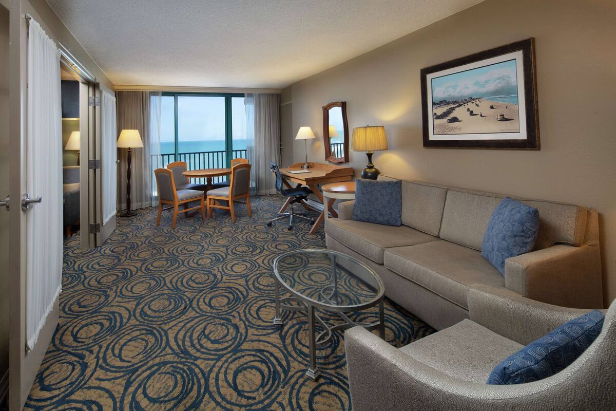 Quarto de hotel com uma cama grande, um sofá, uma escrivaninha e uma cadeira. Há uma janela ampla com vista para a praia.