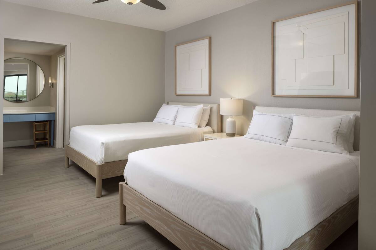 Quarto de hotel com duas camas, uma escrivaninha, uma cadeira, um espelho e obras de arte nas paredes.