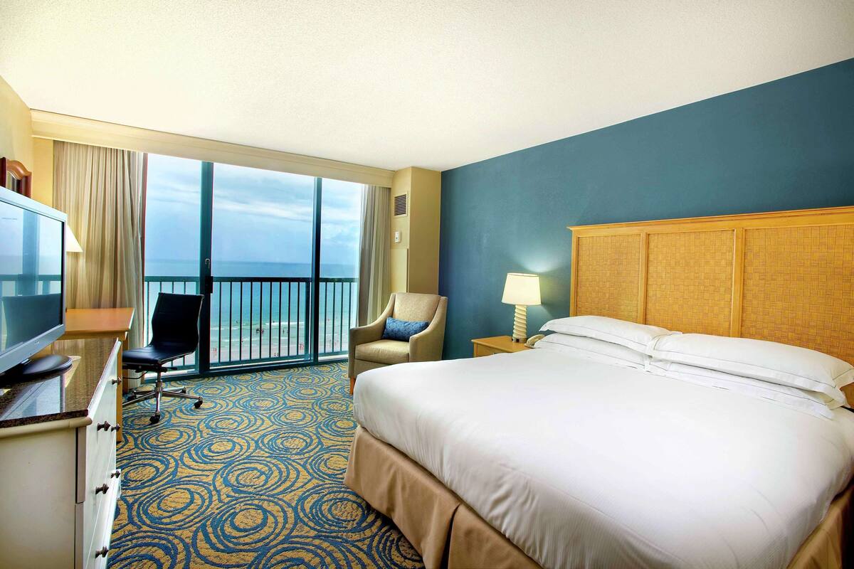 Quarto de hotel com uma cama grande, uma escrivaninha com cadeira, uma televisão e vista para o oceano pelas janelas.