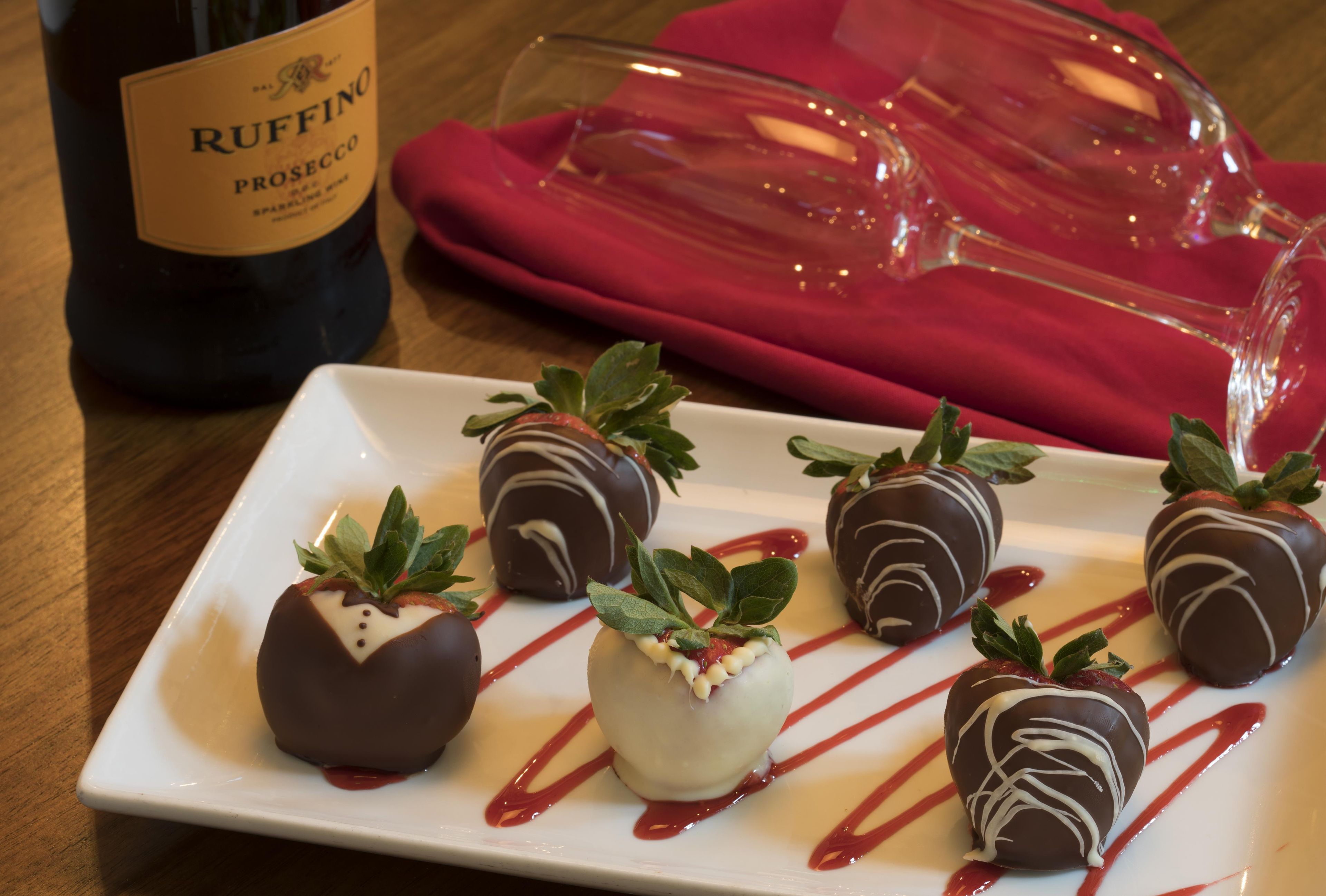 Um prato de morangos cobertos de chocolate com rega de chocolate branco e uma garrafa de vinho ao fundo.