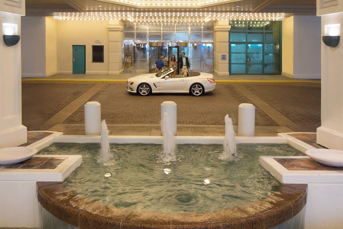 Um saguão de hotel com uma fonte, um carro conversível branco e uma entrada bem iluminada.