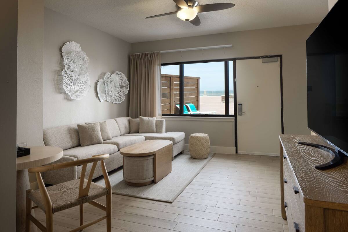 Sala de estar moderna com sofá seccional, mesa de centro redonda, rack para TV e uma grande janela com vista para a praia.