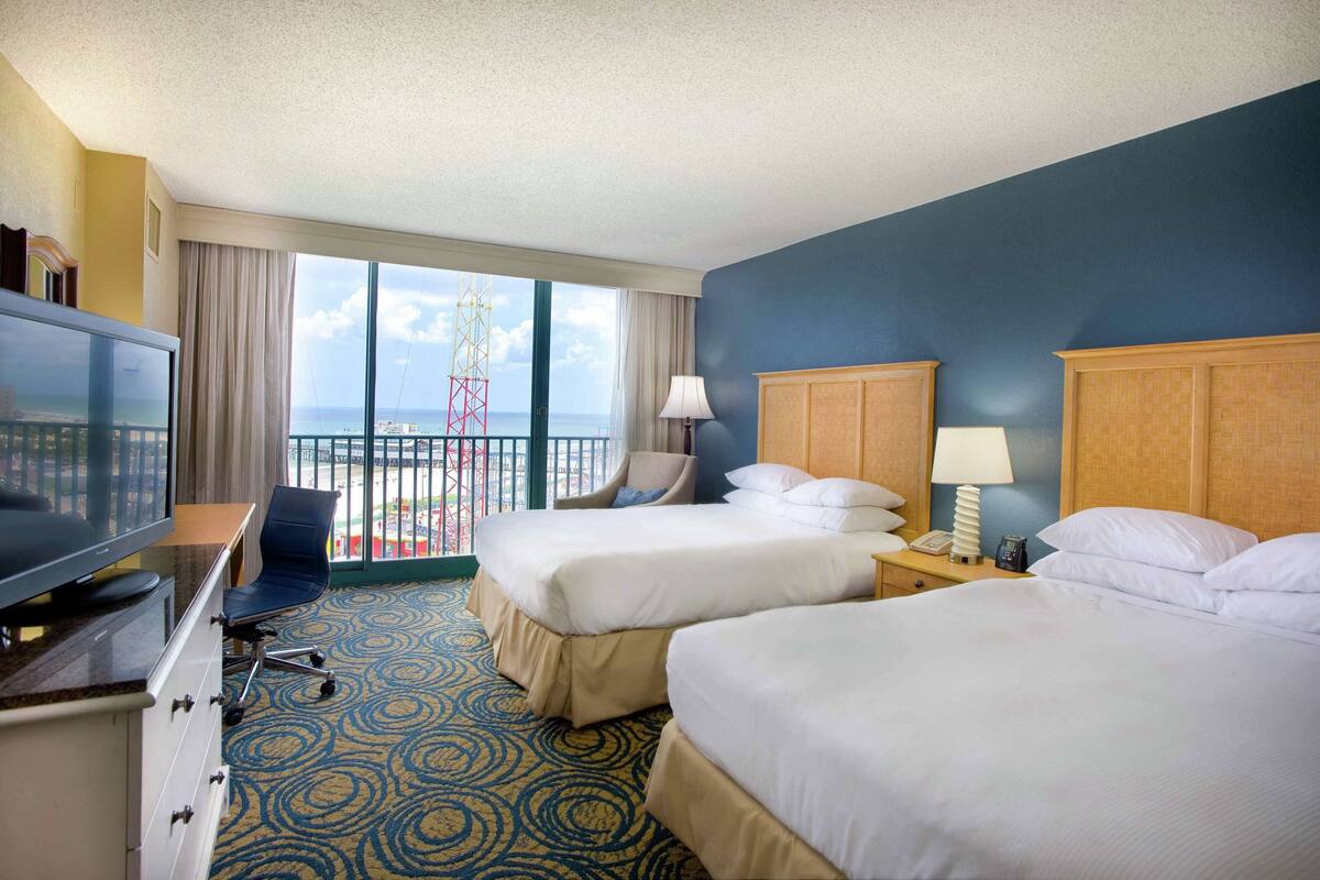 Quarto de hotel com duas camas, uma escrivaninha com televisão, uma cadeira, um abajur e vista para o mar.