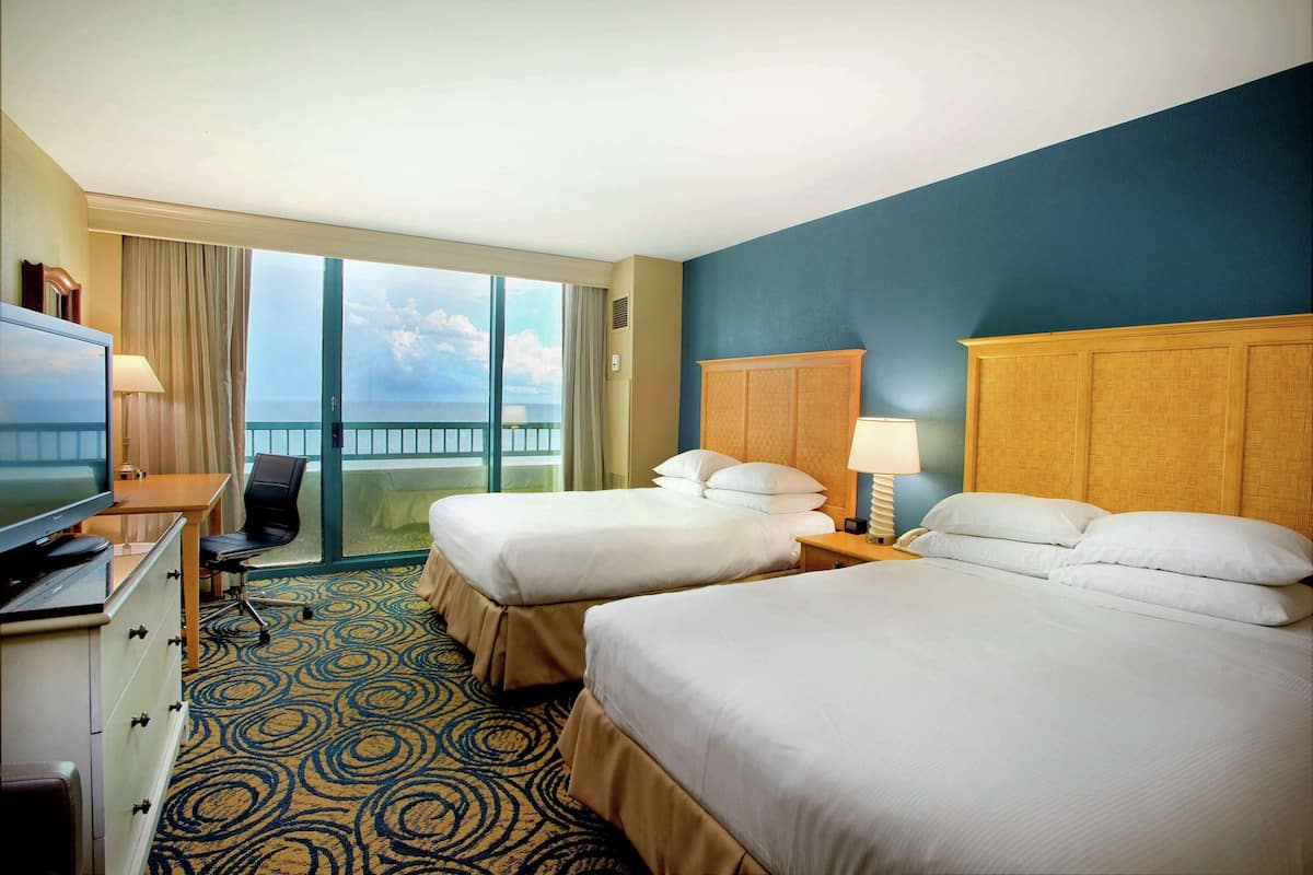 Quarto de hotel com duas camas, uma escrivaninha com televisão, uma cadeira e vista para o oceano.