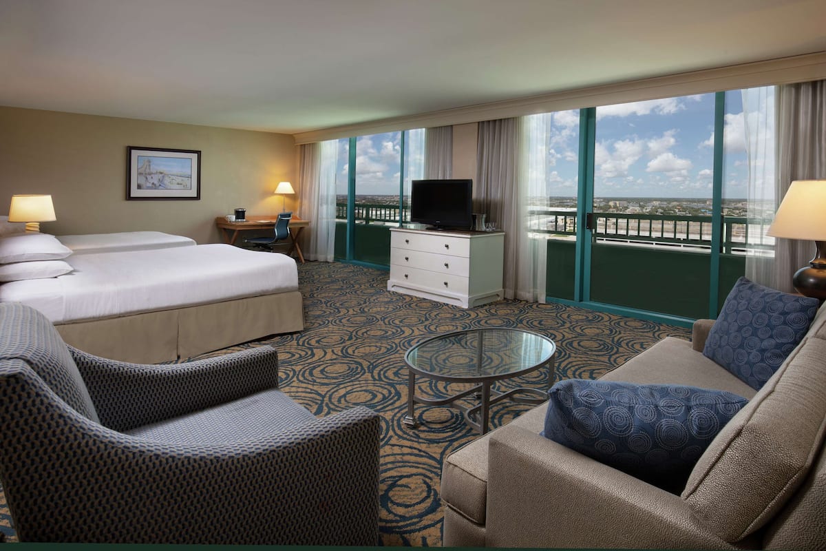 Quarto de hotel com uma cama grande, duas mesas de cabeceira, uma TV, um sofá e uma varanda vista para a cidade.