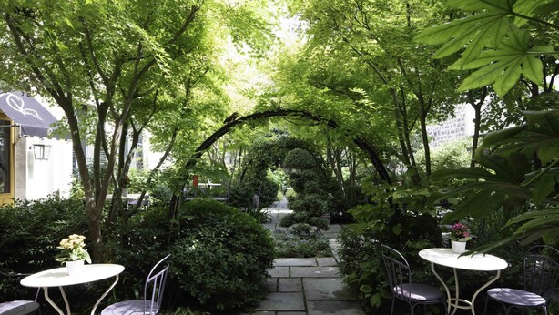 Um jardim externo com um caminho de pedra, uma mesa branca com cadeiras e vegetação exuberante.