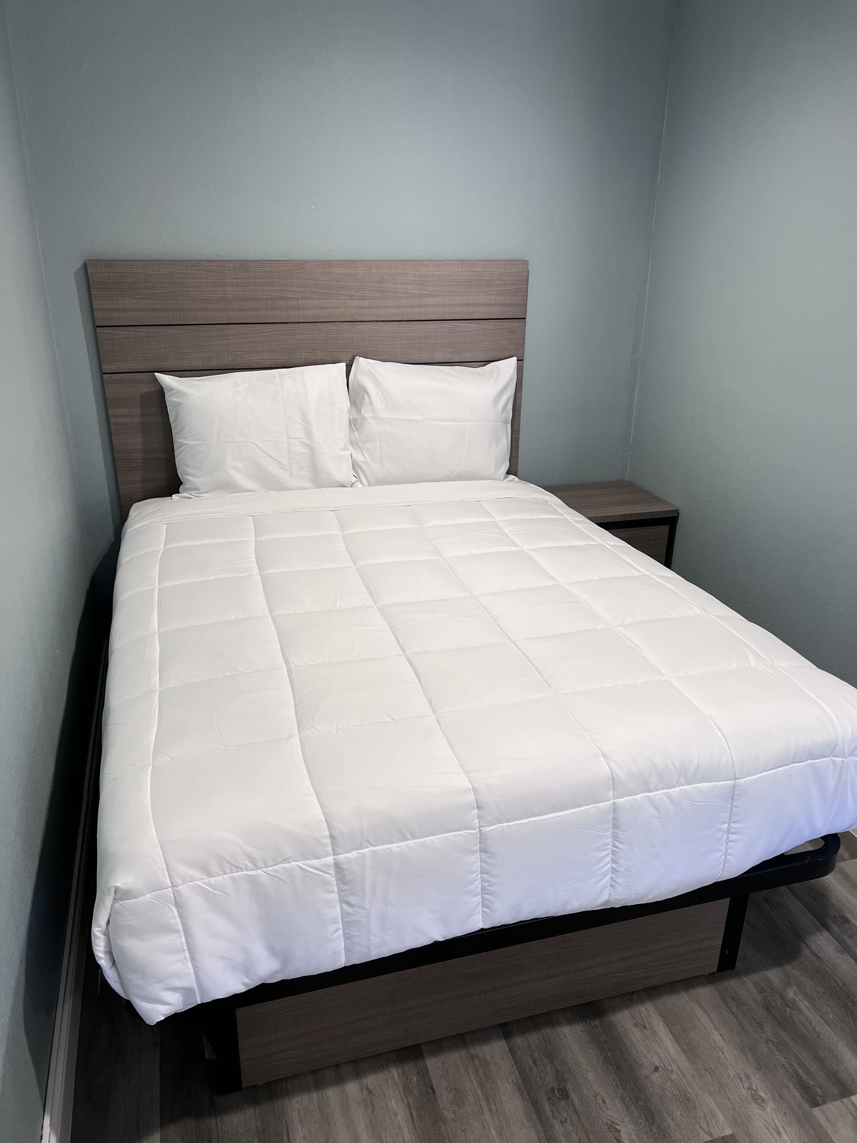Uma cama bem arrumada com um edredom branco acolchoado, uma cabeceira de madeira e uma pequena mesa de cabeceira.