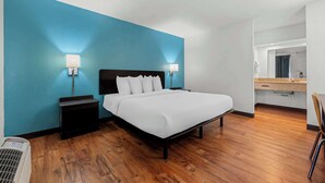 Quarto de hotel moderno com uma cama grande, duas luminárias de cabeceira, uma mesinha de cabeceira, piso de madeira e um banheiro visível através de uma porta aberta.