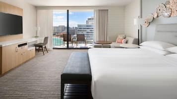 Quarto de hotel com uma cama grande, uma mesa de trabalho com uma TV, uma varanda com cadeiras e vista para a cidade.