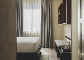 Quarto de hotel com cama, um banco, uma pequena mesa com produtos de higiene e um guarda-roupa.