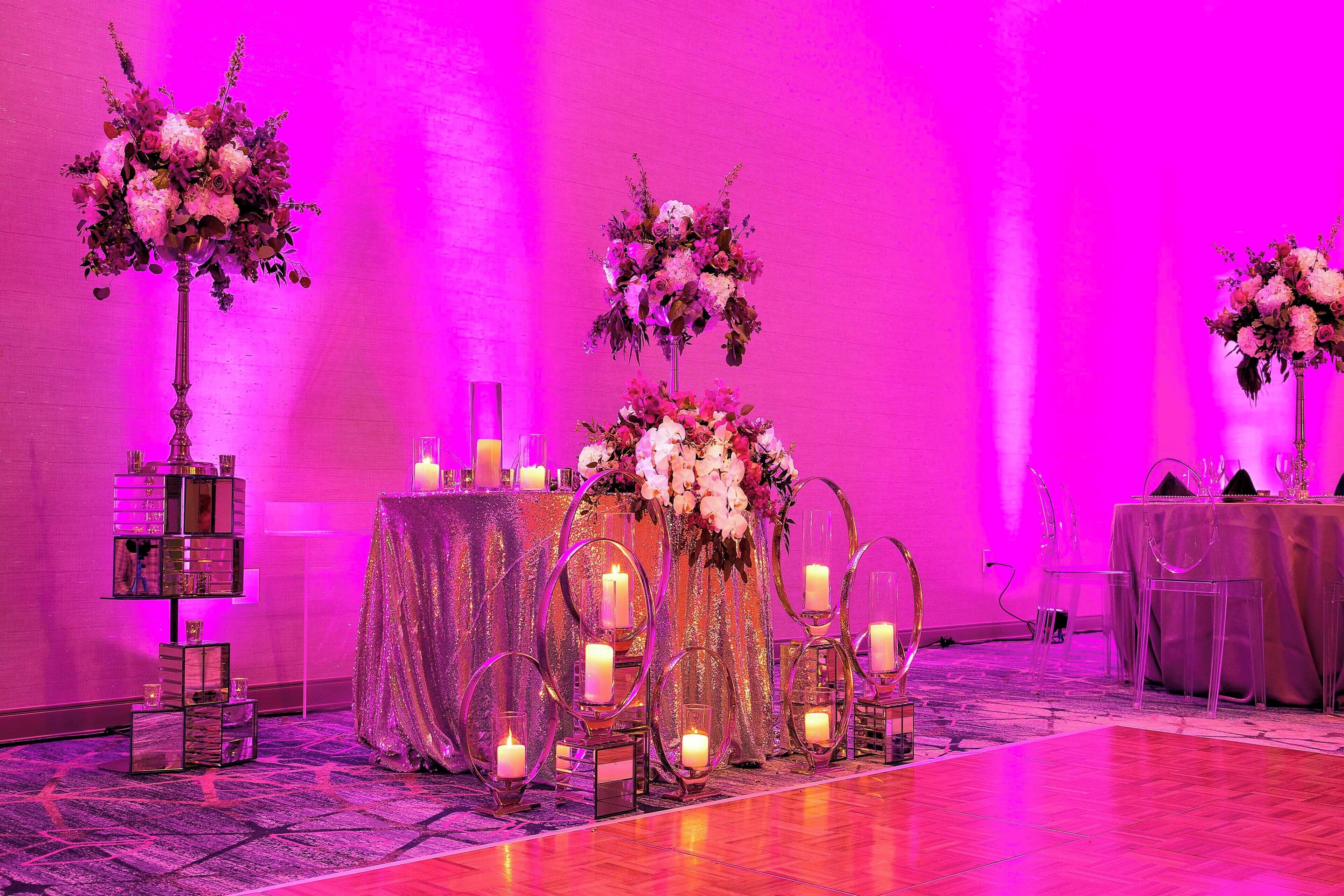 Uma área de recepção com uma mesa decorada com flores e velas, rodeada por suportes de vidro transparente para velas.