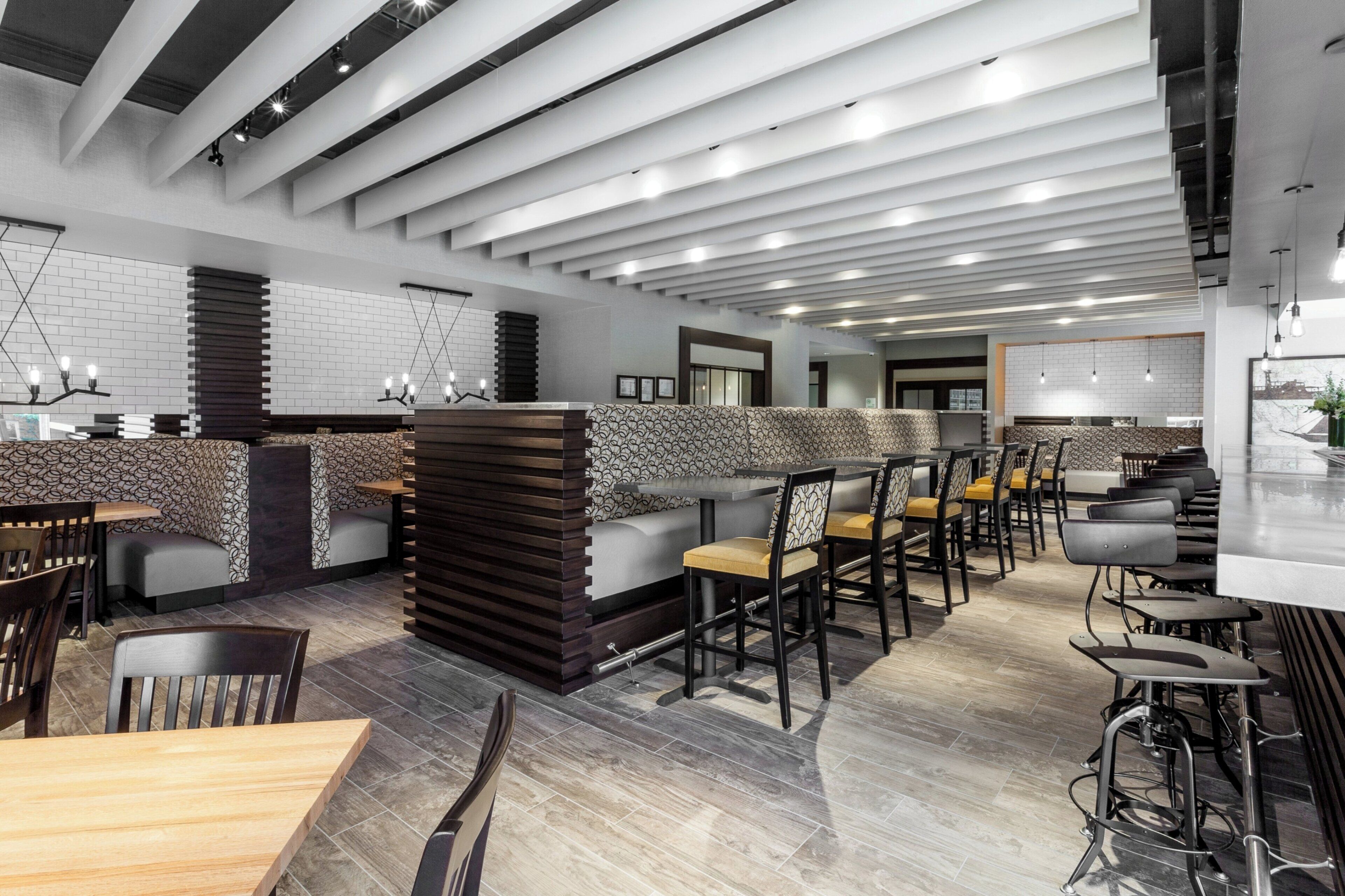 Interior moderno de restaurante com pé-direito alto, vigas aparentes e variedade de assentos, incluindo cabines e mesas.