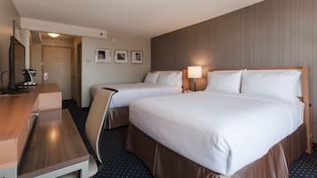 Quarto de hotel com duas camas, uma mesa, uma cadeira, luminária de mesa e televisão fixada na parede.