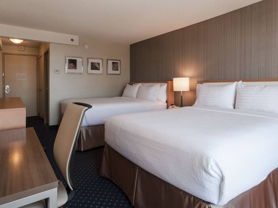 Quarto de hotel com duas camas, uma mesa, uma cadeira, luminária de mesa e televisão fixada na parede.