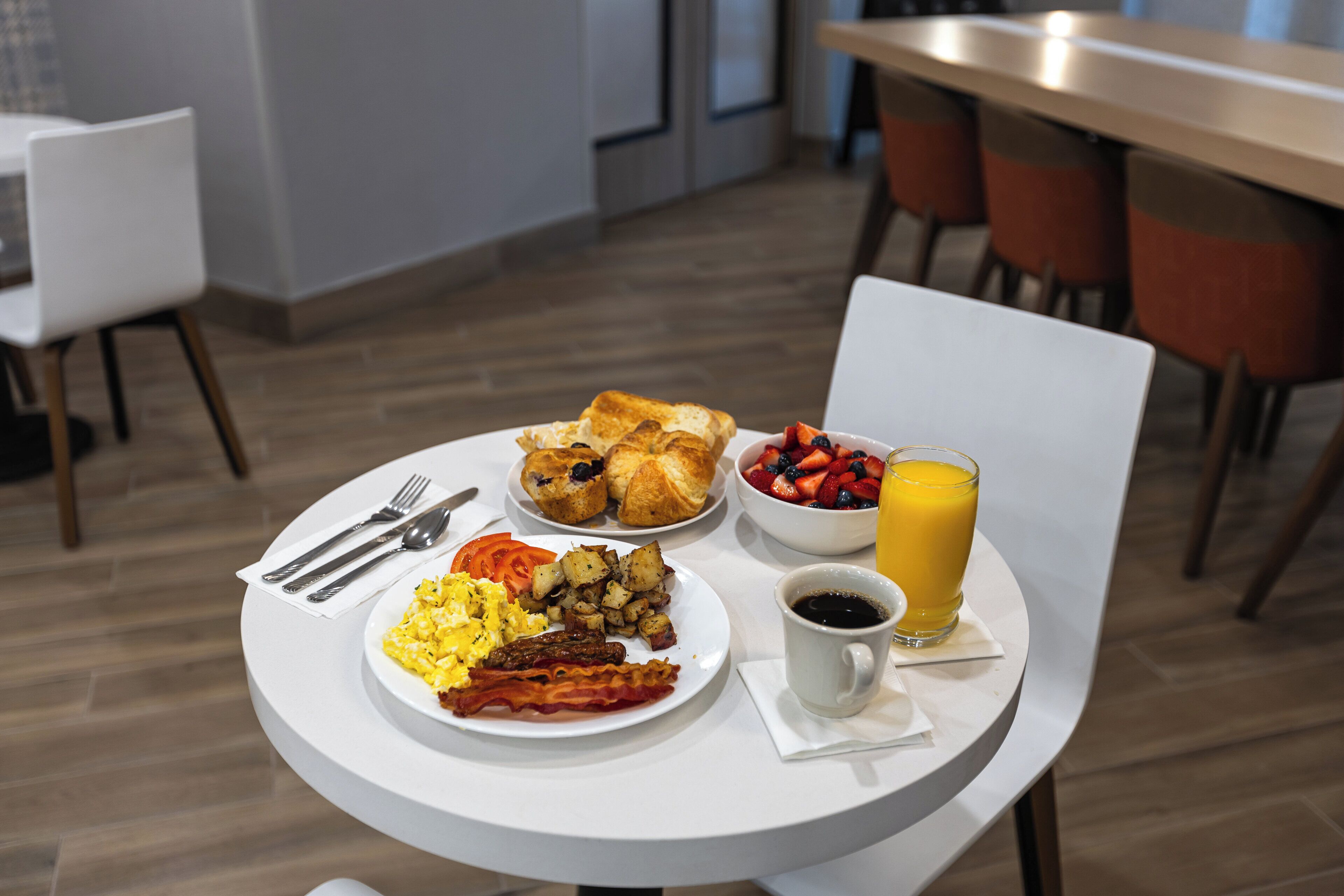 Uma mesa de café da manhã com pratos de ovos, bacon, torradas e frutas, um copo de suco de laranja e uma xícara de café.