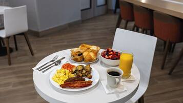 Uma mesa de café da manhã com pratos de ovos, bacon, torradas e frutas, um copo de suco de laranja e uma xícara de café.