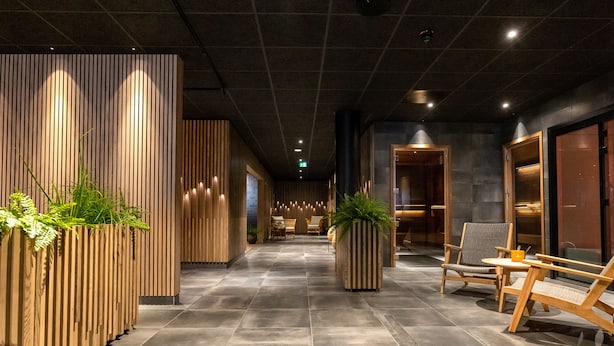 Um saguão moderno de hotel com balcão de atendimento em madeira, área de estar e uma grande planta em vaso.