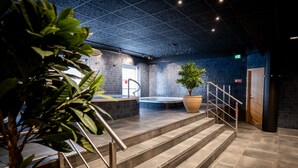 Uma área interna de piscina moderna, com uma escadaria que leva a uma banheira de hidromassagem, cercada por paredes de azulejos escuros e plantas em vasos.