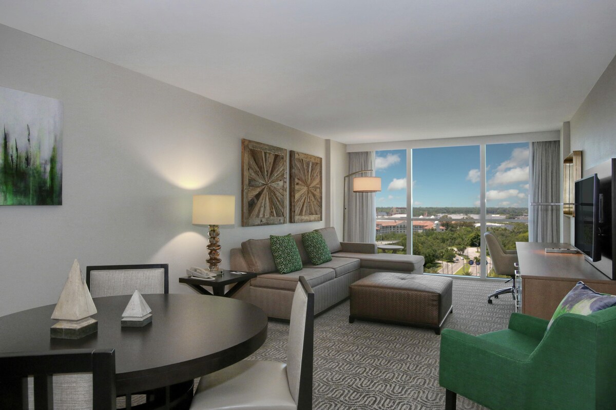 Quarto de hotel moderno com área de refeições, sofá, poltrona verde, televisão e grandes janelas com vista para a cidade.