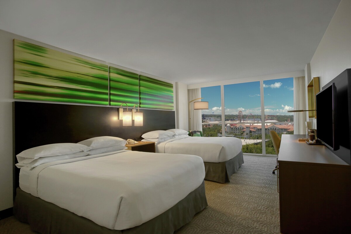 Quarto de hotel com duas camas, uma janela grande com vista para a cidade, uma televisão de tela plana e uma pintura abstrata moderna na parede.