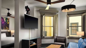 Quarto de hotel moderno com cama, televisão, sofá, mesa de centro e ventoinha no teto.
