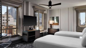 Quarto de hotel com duas camas, televisão, mesa de trabalho com candeeiro e amplas janelas com persianas.