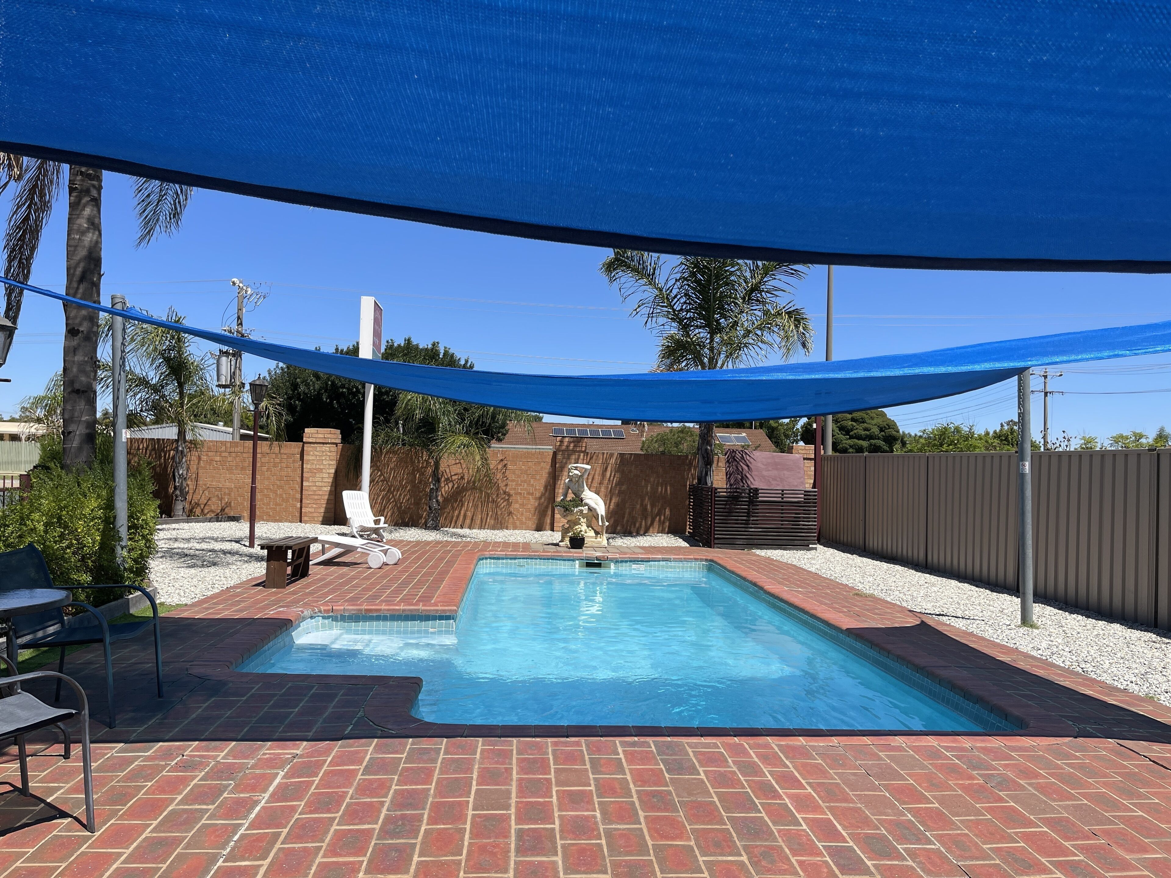 Piscina retangular com deck de tijolos, rodeada por uma cobertura azul de tecido, uma cadeira branca e uma pequena mesa.