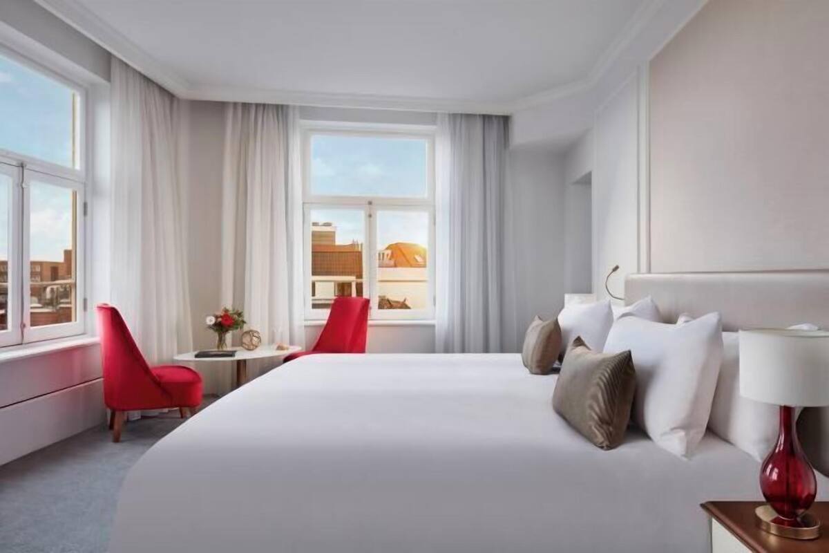 Quarto de hotel com uma cama grande, uma cadeira vermelha, uma mesinha com um vaso e vista para edifícios.