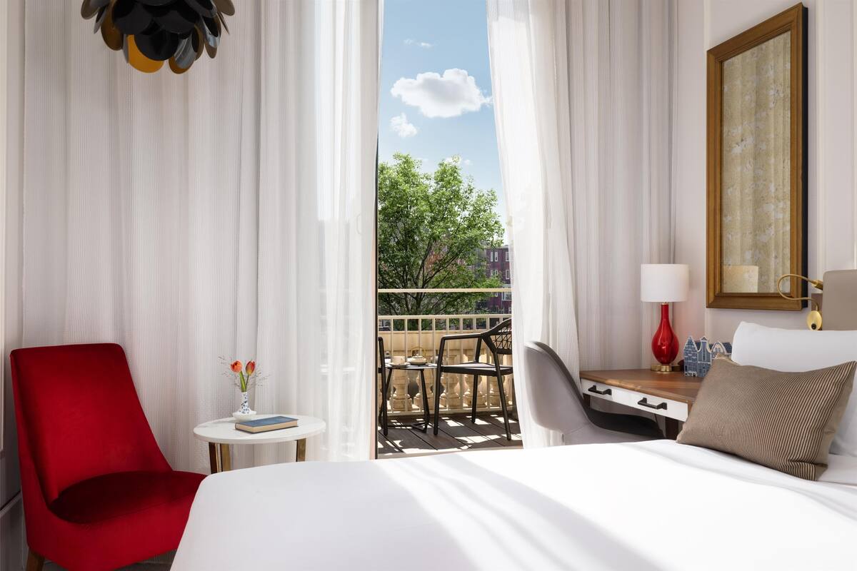 Quarto de hotel com uma cama, uma cadeira vermelha, uma mesinha com um vaso de flores, uma escrivaninha com abajur e vista para o exterior.