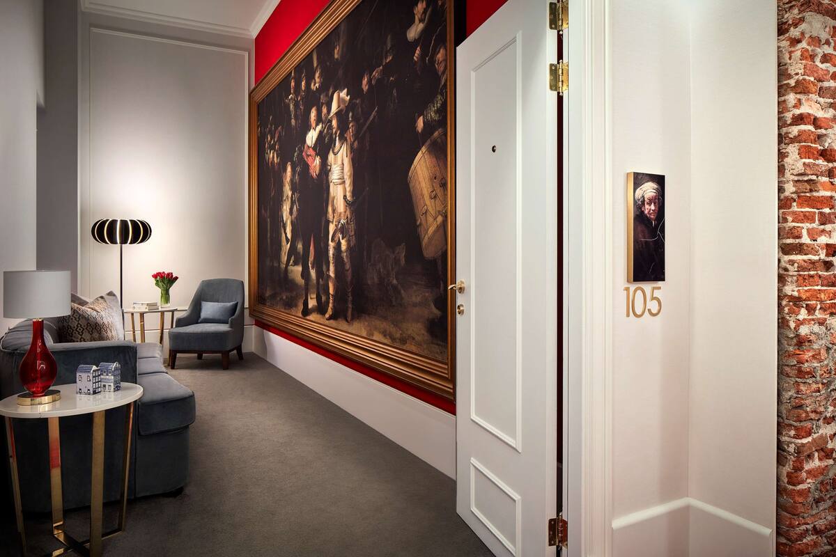 Corredor com um quadro clássico grande, uma luminária moderna, um sofá e uma porta com o número 105.