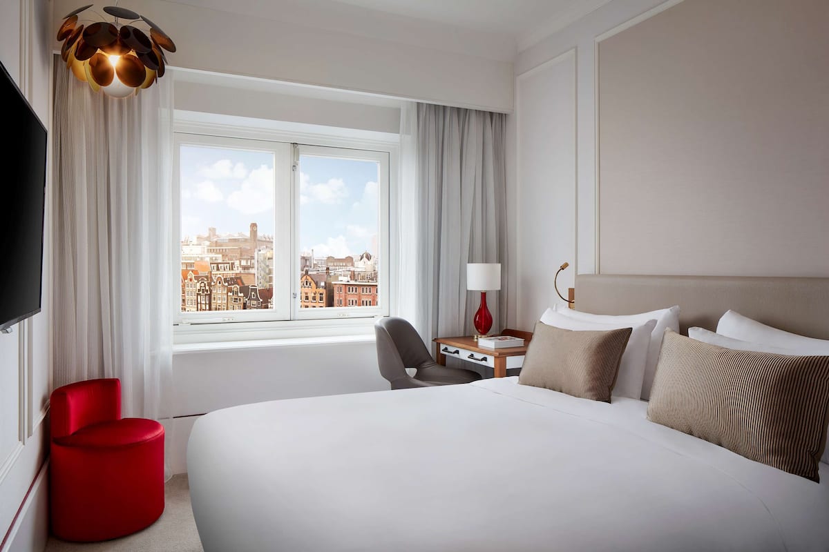 Quarto de hotel com uma cama grande, uma cadeira vermelha, uma escrivaninha com abajur e vista para a cidade através da janela.
