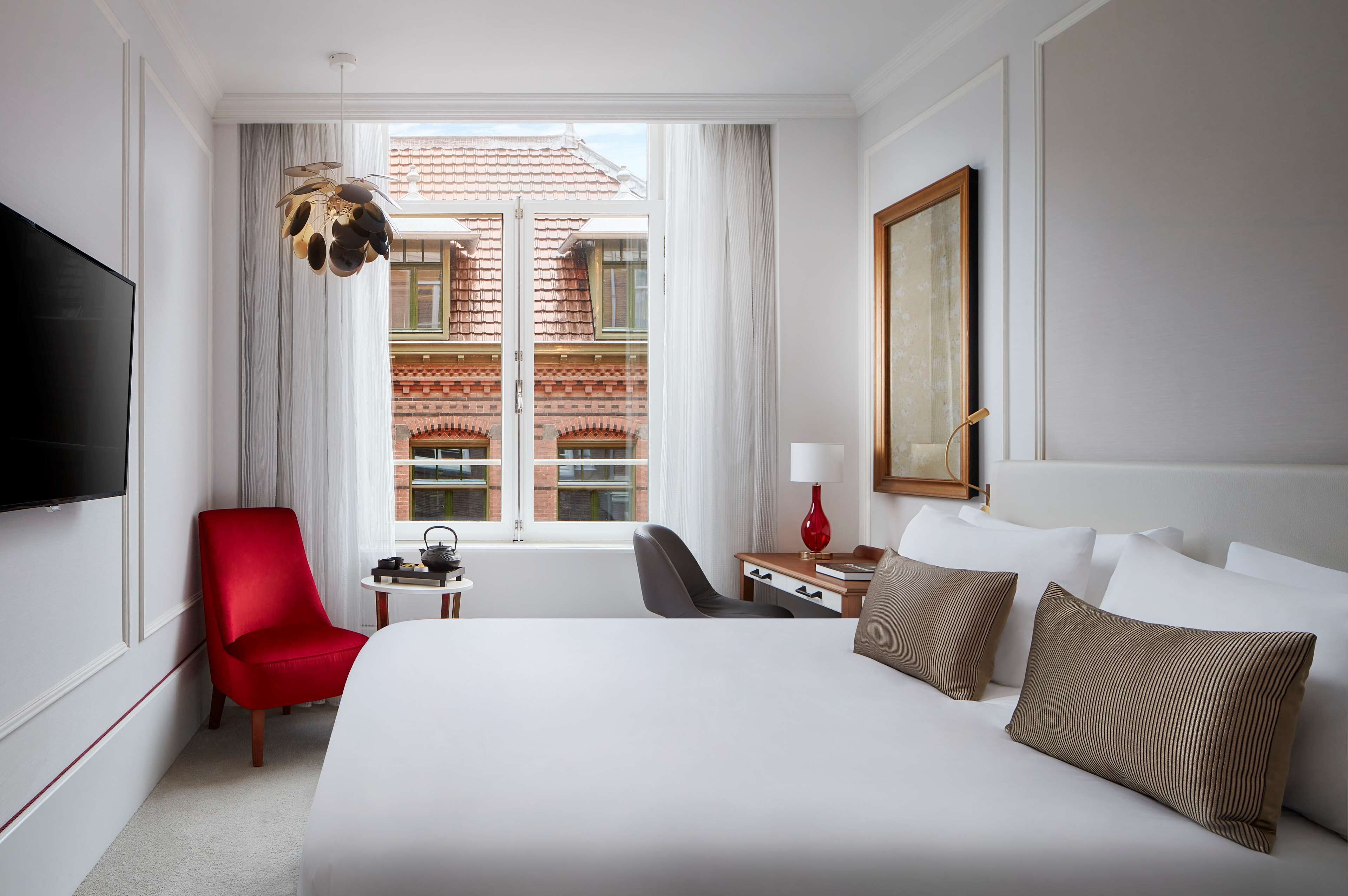Quarto de hotel com cama, televisão, uma cadeira vermelha, mesa pequena com abajur e janela com vista para um edifício.